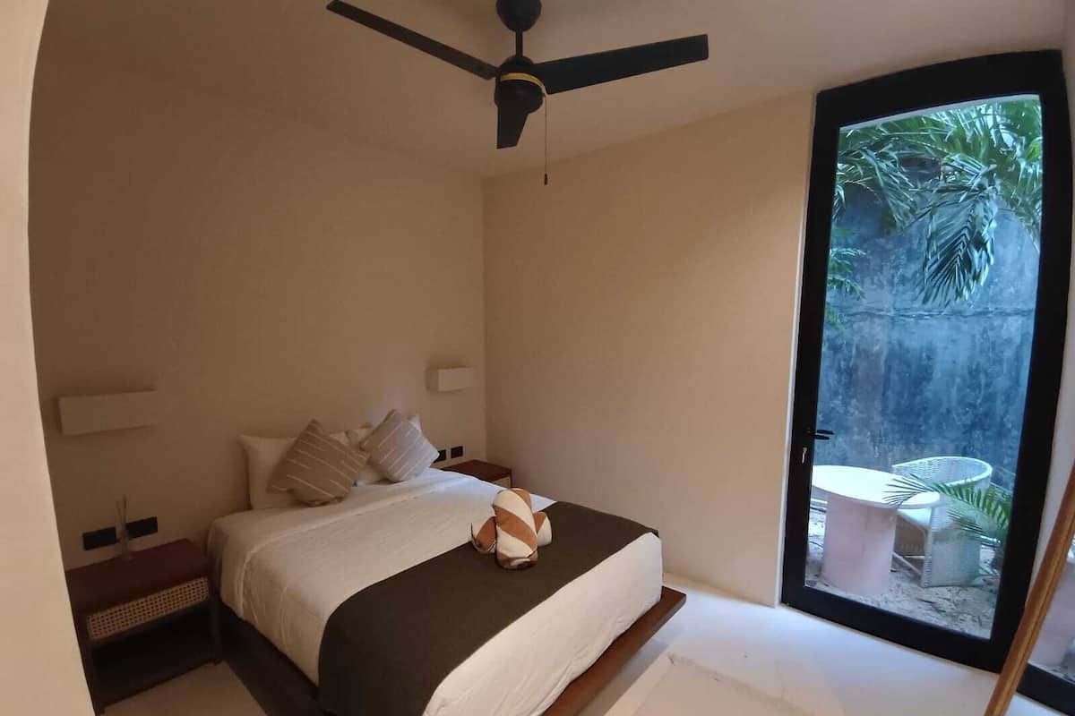 Quarto com cama, ventoinha no teto, mesa de cabeceira e vista para um pátio com mesa e cadeiras.
