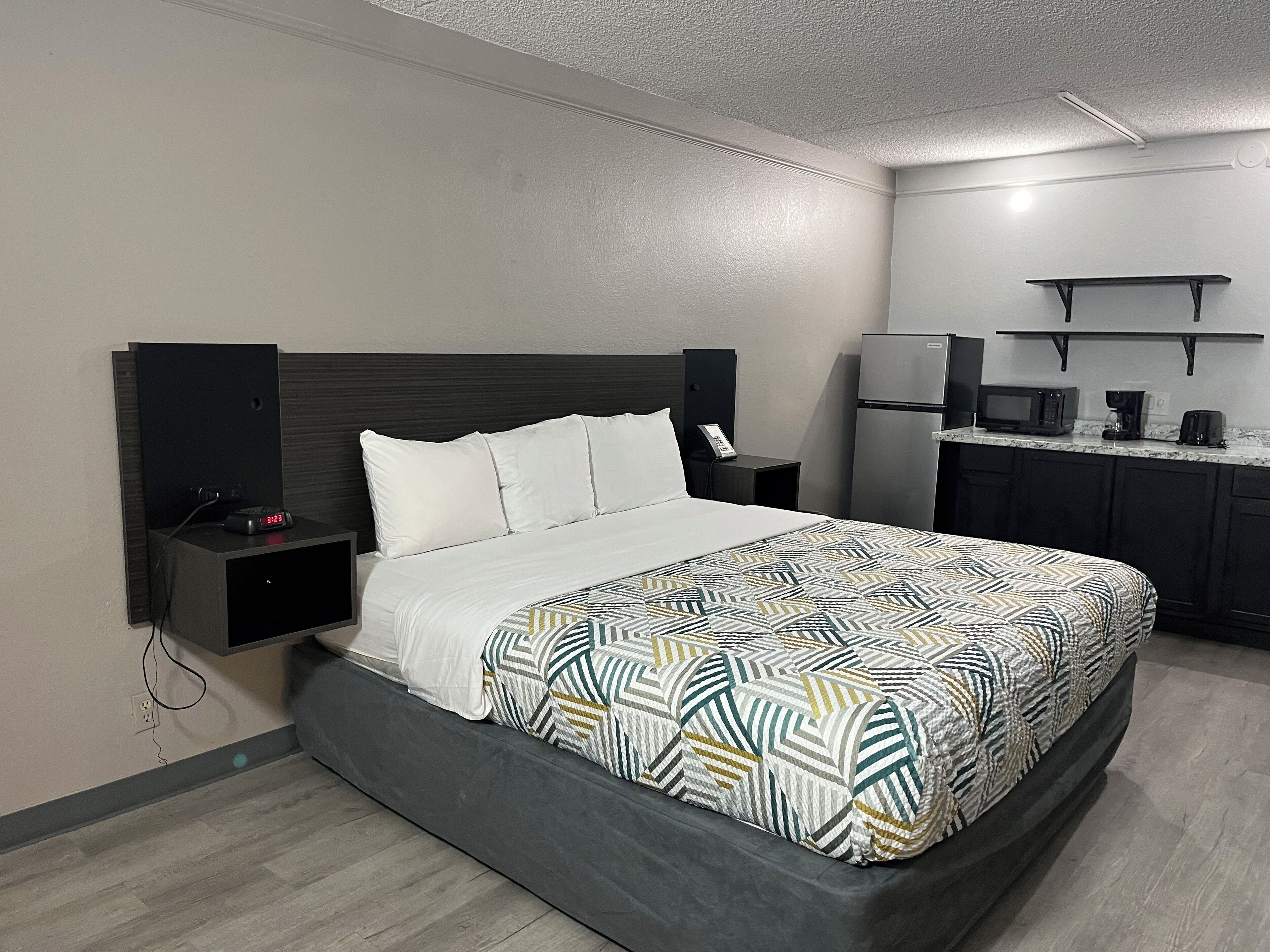 Quarto de hotel moderno com cama, mesa de cabeceira, escrivaninha, micro-ondas, frigorífico e máquina de café.