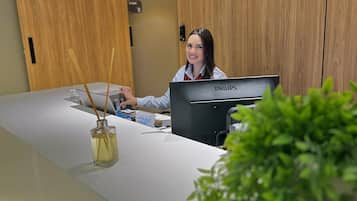 Bancada de recepção com um computador, um frasco de perfume e uma planta em primeiro plano.