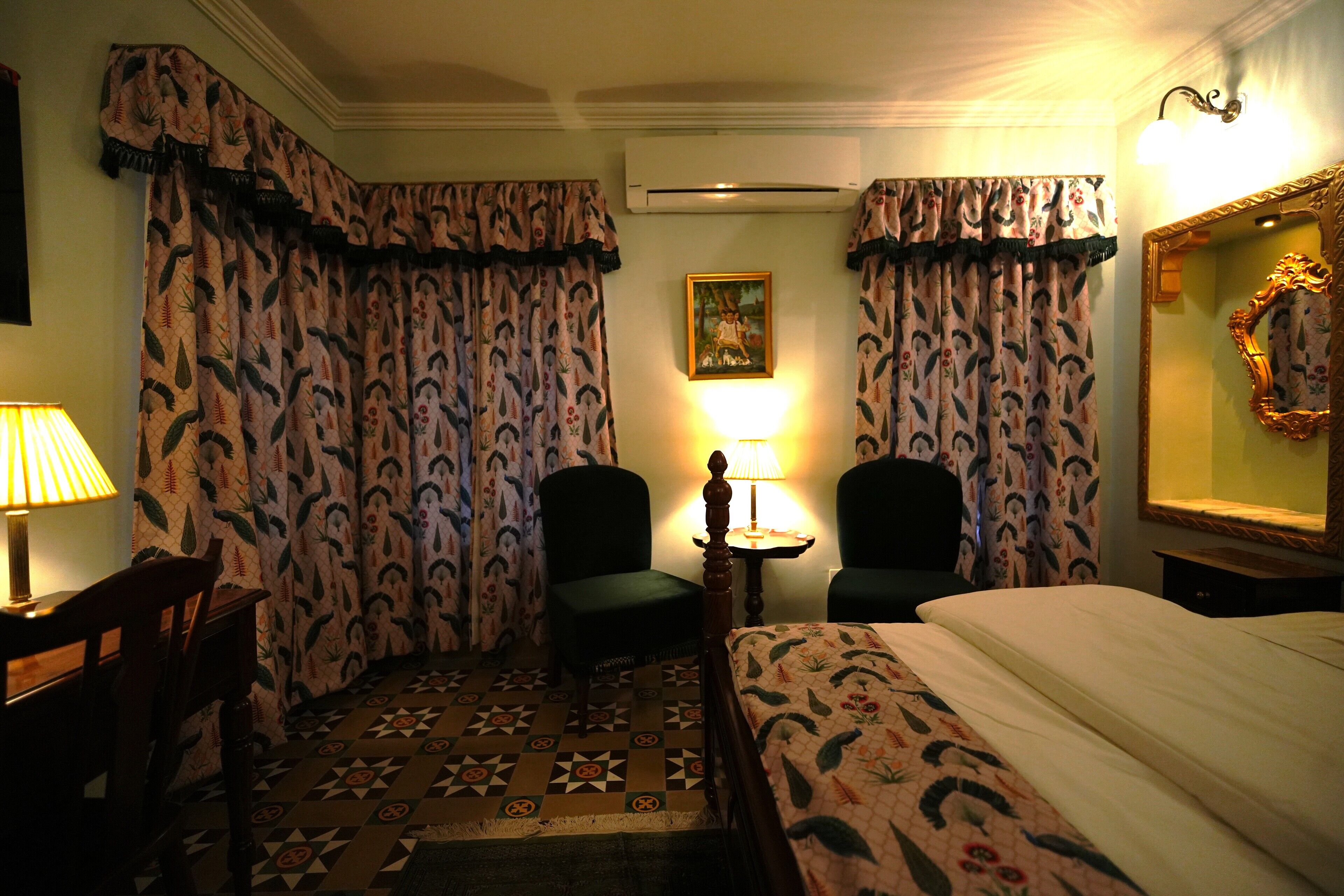 Quarto de hotel com cama, duas cadeiras, mesa com abajur, cortinas estampadas, quadro na parede e ar condicionado.