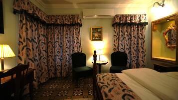 Quarto de hotel com cama, duas cadeiras, mesa com abajur, cortinas estampadas, quadro na parede e ar condicionado.