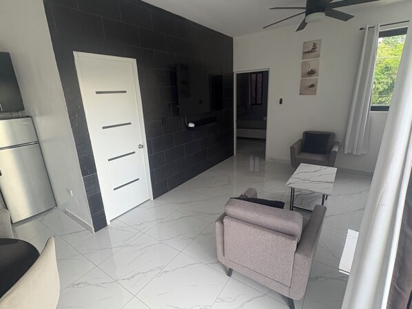Sala de estar moderna com sofá, mesa de centro e televisão. Possui área de cozinha com geladeira e parede revestida de azulejos pretos. É visível um ventilador de teto e uma janela com cortinas.