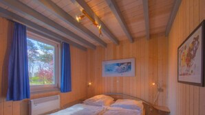 Quarto de cabana de madeira com cama, janela com cortinas, radiador e um quadro na parede.