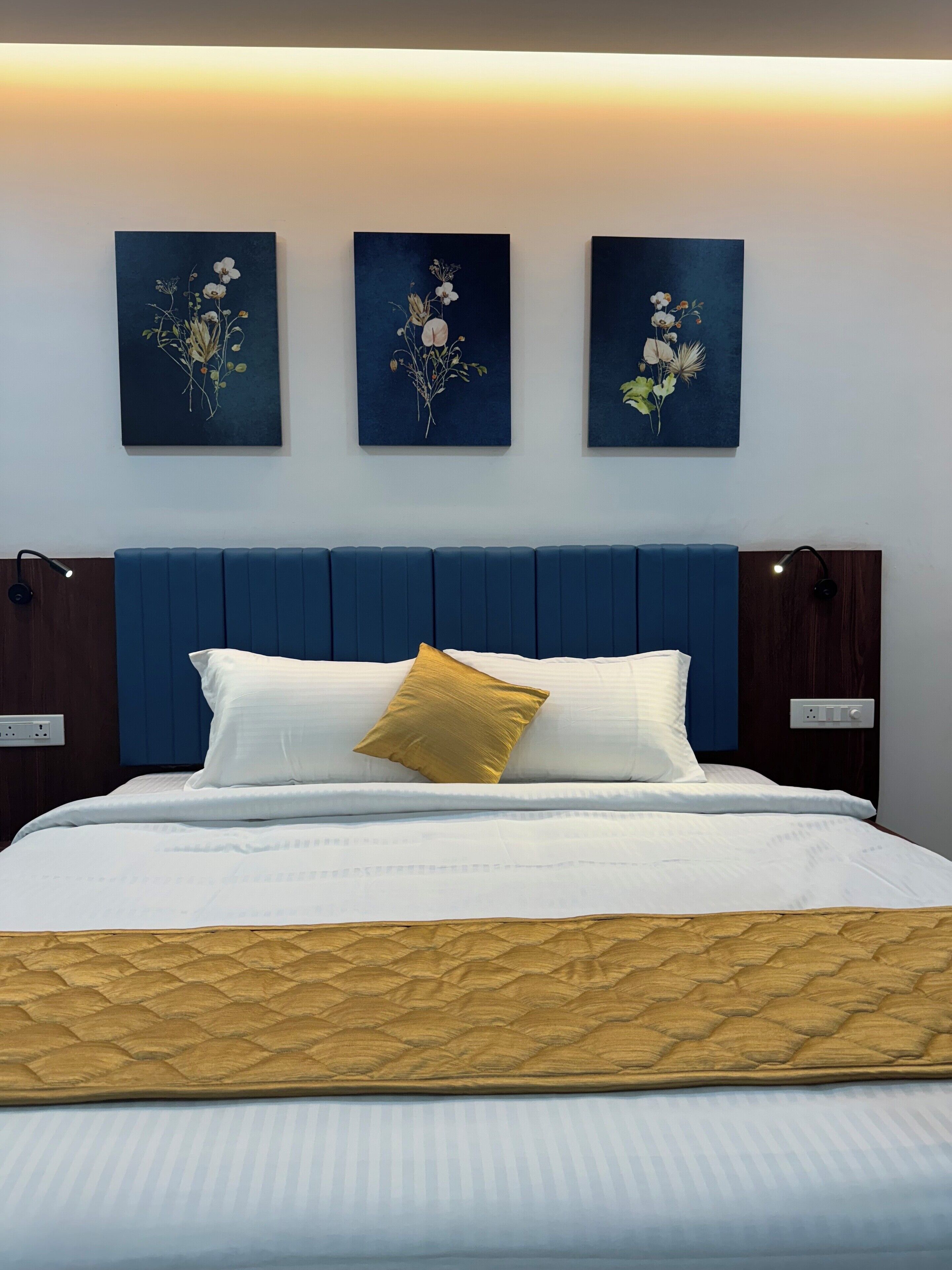 Uma cama com roupa de cama branca e dourada, uma cabeceira azul e três quadros florais na parede.