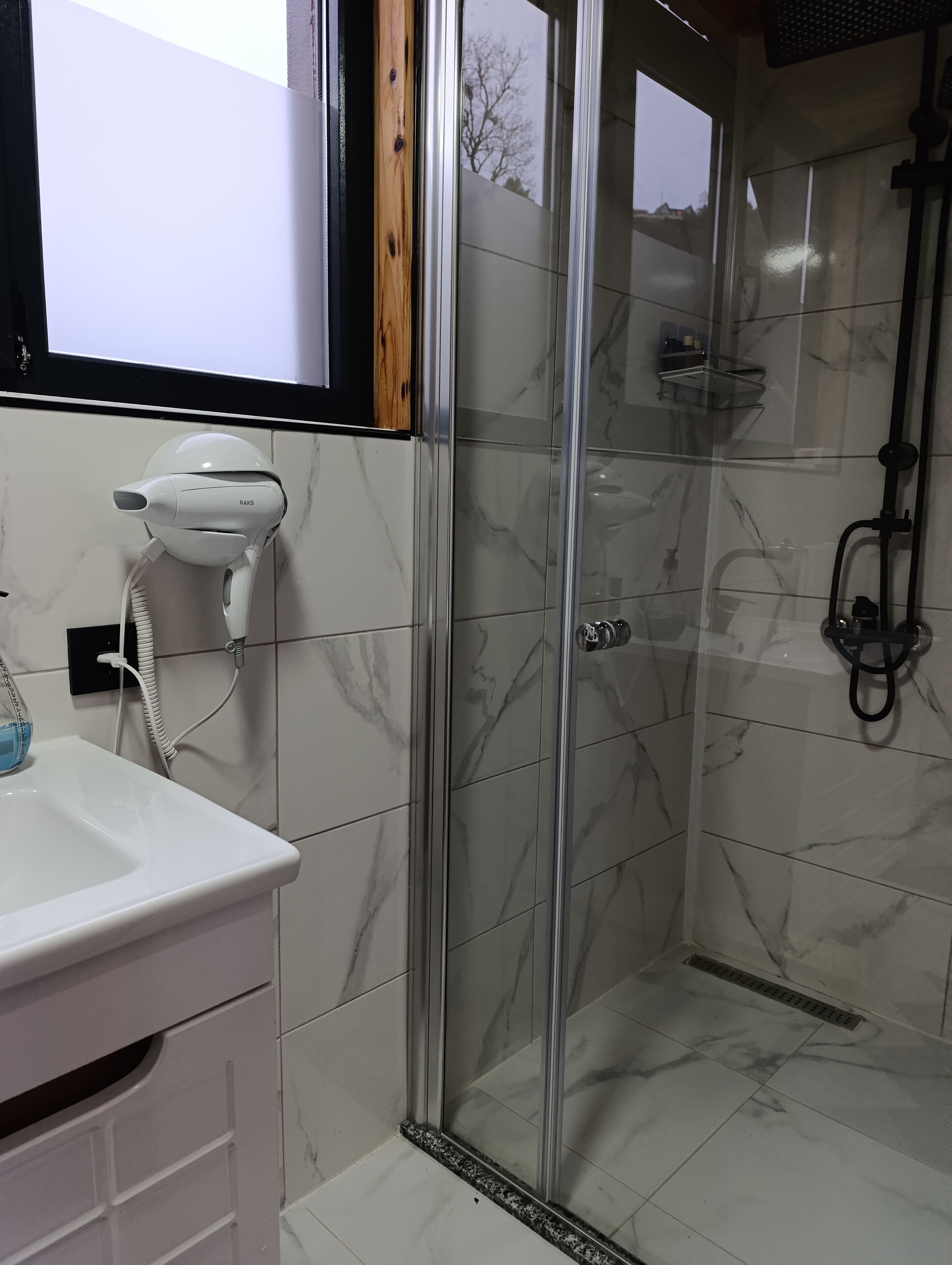 Casa de banho com cabine de chuveiro em vidro, pia branca e secador de cabelo fixado na parede.
