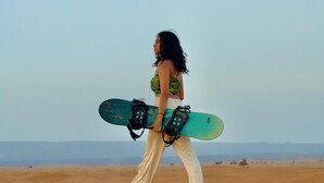 Uma pessoa caminhando no deserto com uma prancha de snowboard.