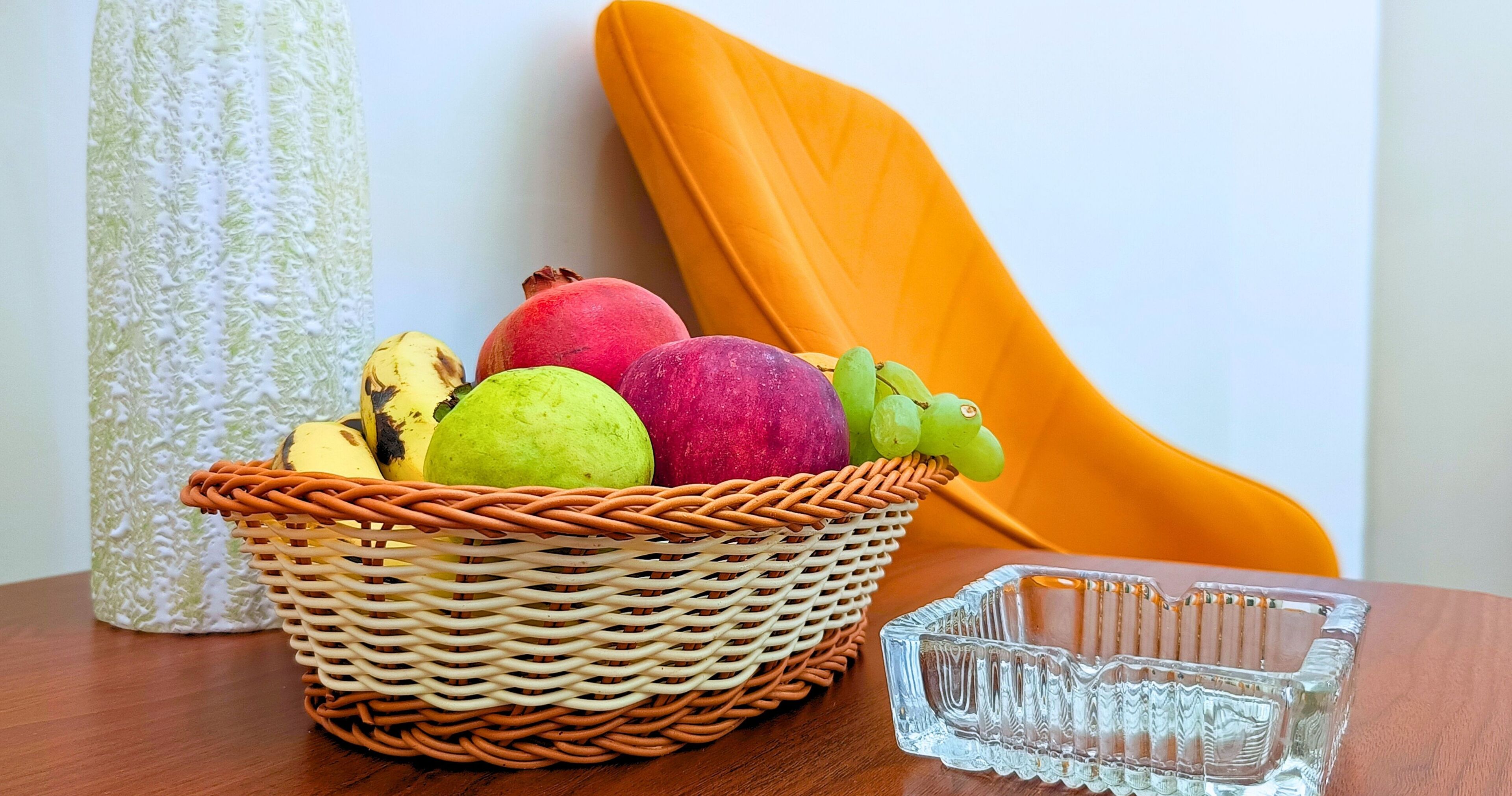 Uma cesta de vime cheia de frutas sobre uma mesa de madeira, ao lado de um prato de vidro transparente.