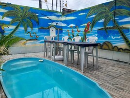 Piscina com um mural, mesa com cadeiras e um deck de madeira.