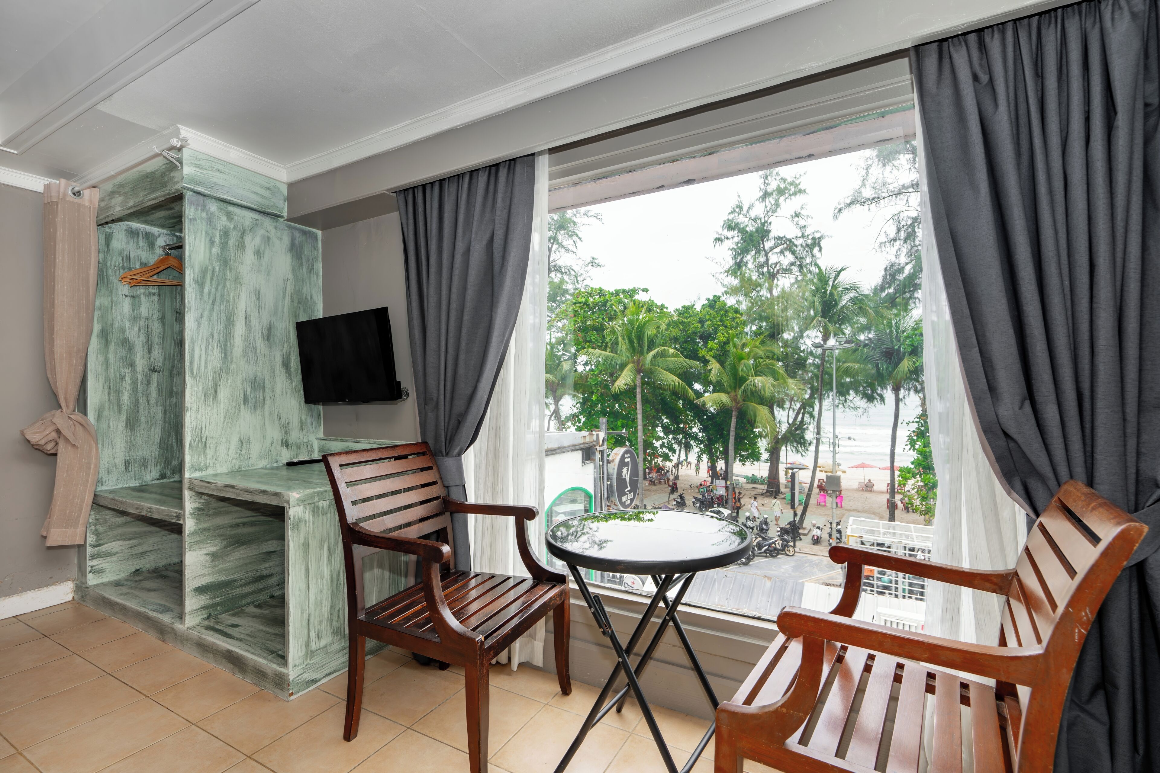 Quarto com um banco de madeira, uma mesa redonda pequena, televisão presa na parede e vista para uma praia com palmeiras.