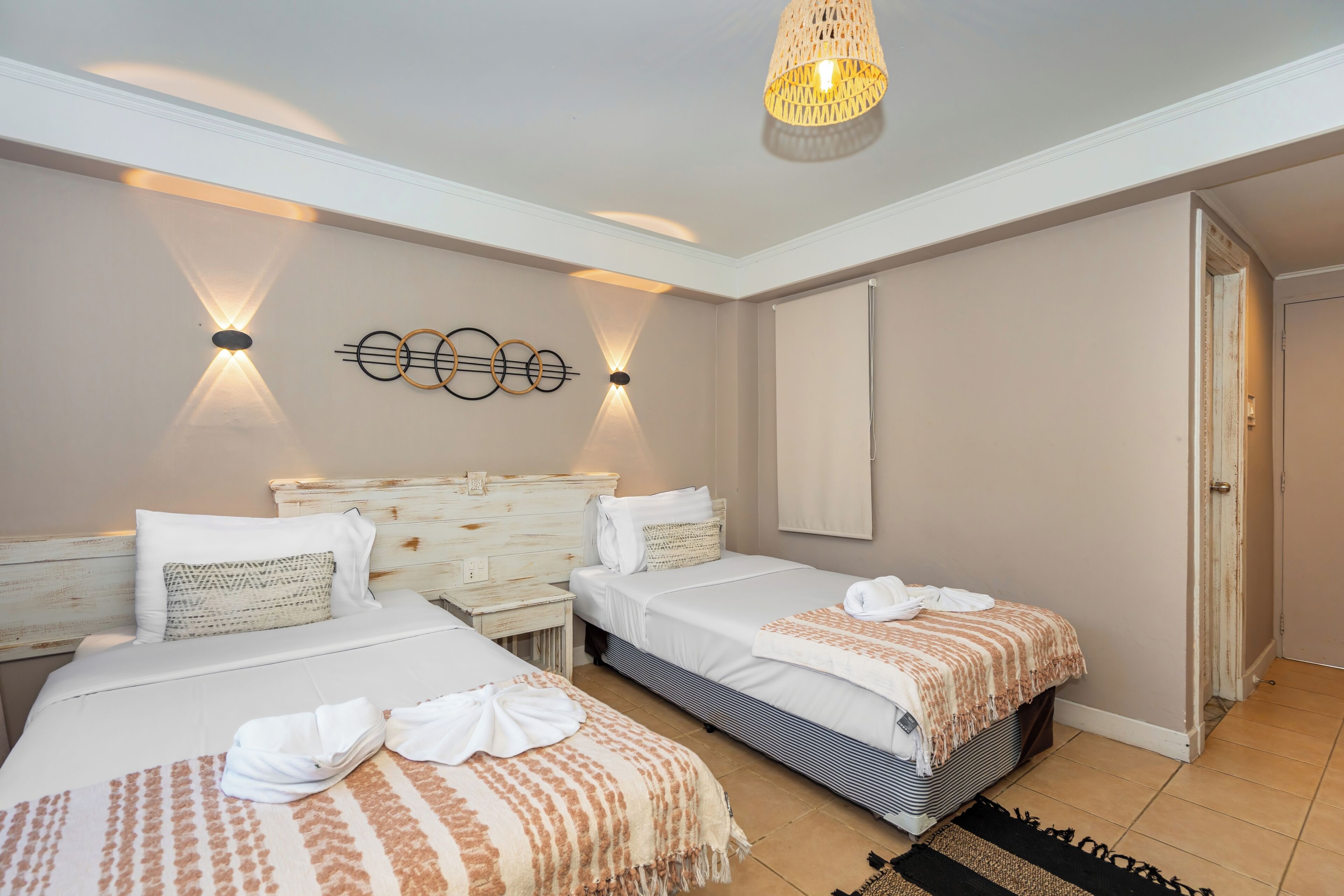 Quarto de hotel com duas camas, cabeceira de madeira, luminárias fixadas na parede e um candeeiro pendente, além de uma porta.