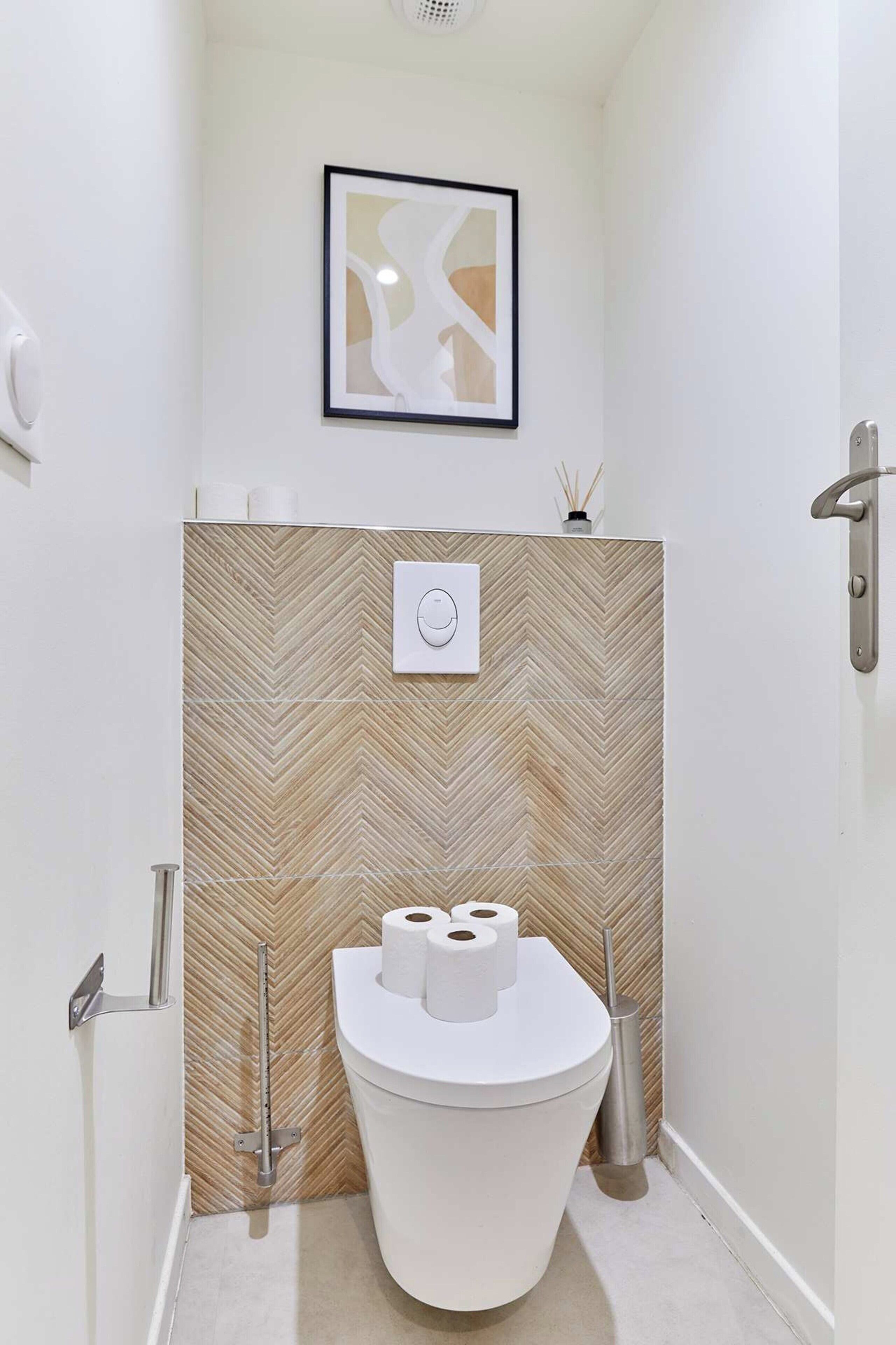 Um banheiro moderno com vaso sanitário branco, parede com padrão herringbone, obra de arte abstrata emoldurada e porta-papel higiênico.
