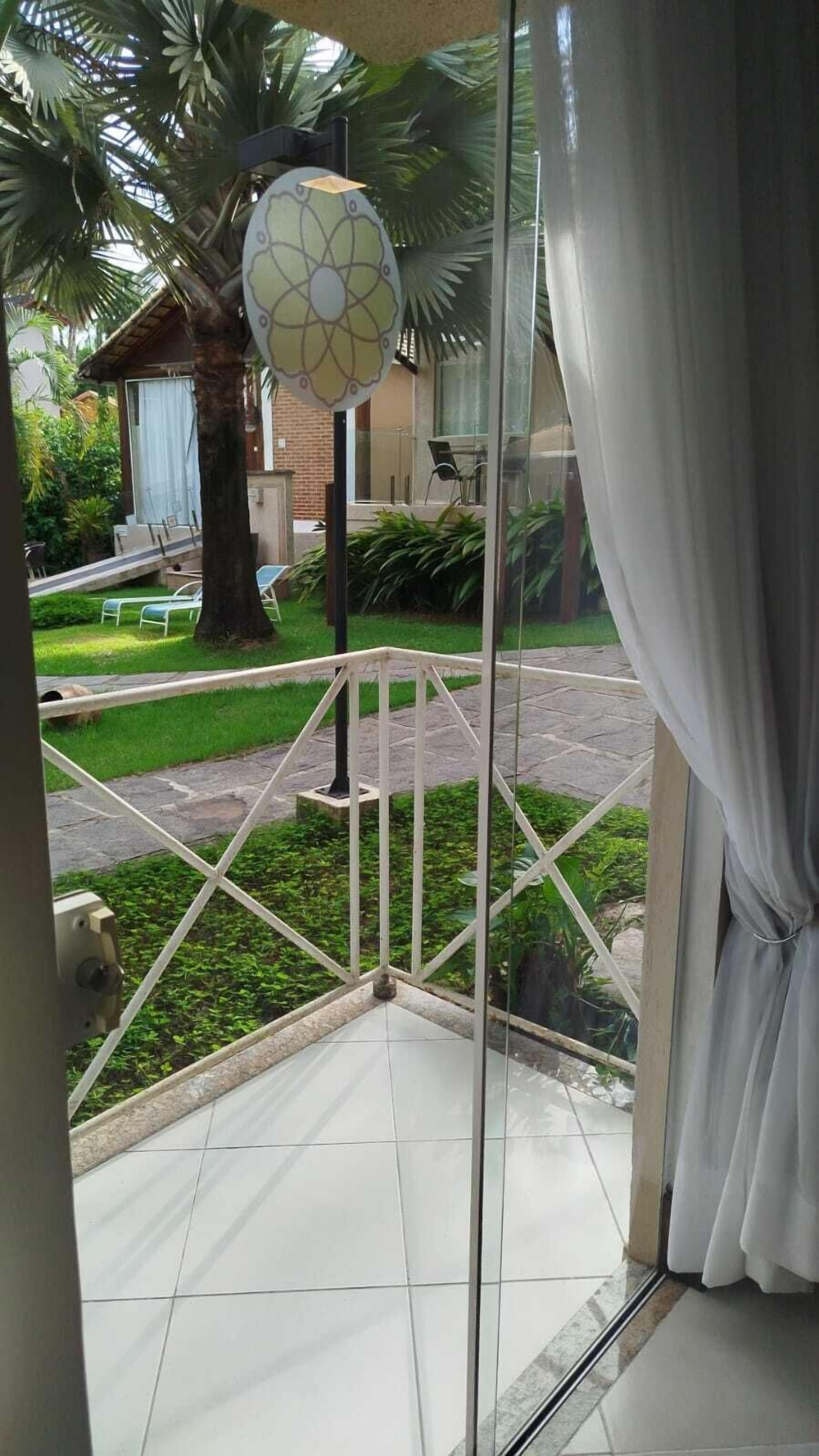 Uma varanda de vidro com guarda-corrimão de metal, uma palmeira e vista para um jardim e um prédio ao fundo.