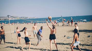 Jogo de voleibol de praia com jogadores de shorts e regatas, rede de voleibol e uma praia de areia com pessoas ao fundo.