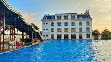 Uma grande piscina com área coberta e um edifício com várias janelas e sacadas ao fundo.