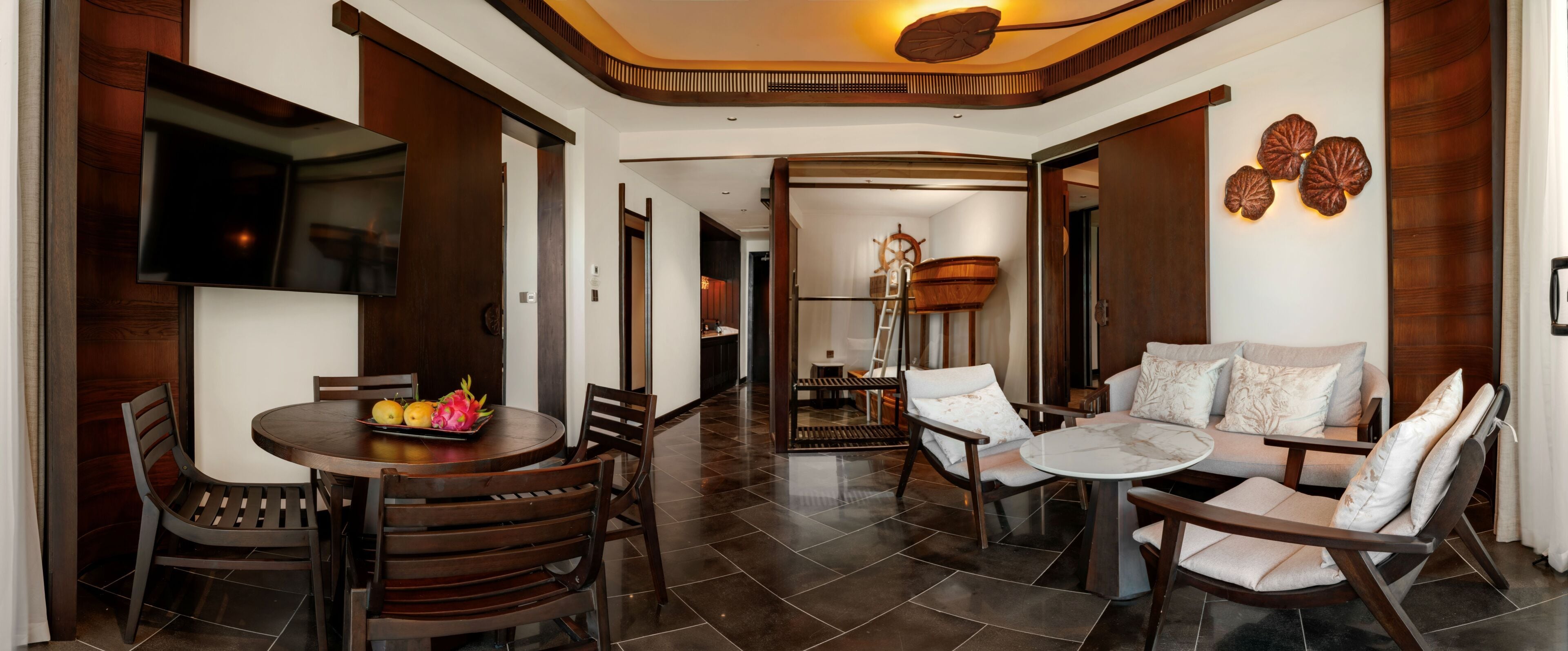 Um lobby de hotel moderno com uma mesa de jantar redonda, cadeiras de madeira, um conjunto de sofás e uma televisão fixada na parede.