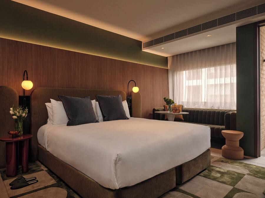 Quarto de hotel moderno com uma cama grande, duas luminárias de cabeceira, uma mesinha com um vaso de flores e vista da cidade por grandes janelas.