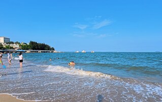 Uma praia com pessoas nadando e caminhando pela orla, céu claro e edifícios ao longe.