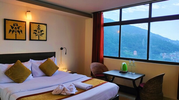 Quarto de hotel com cama, mesa de cabeceira, mesa com chaleira e garrafas, uma cadeira e uma grande janela com vista para as montanhas.