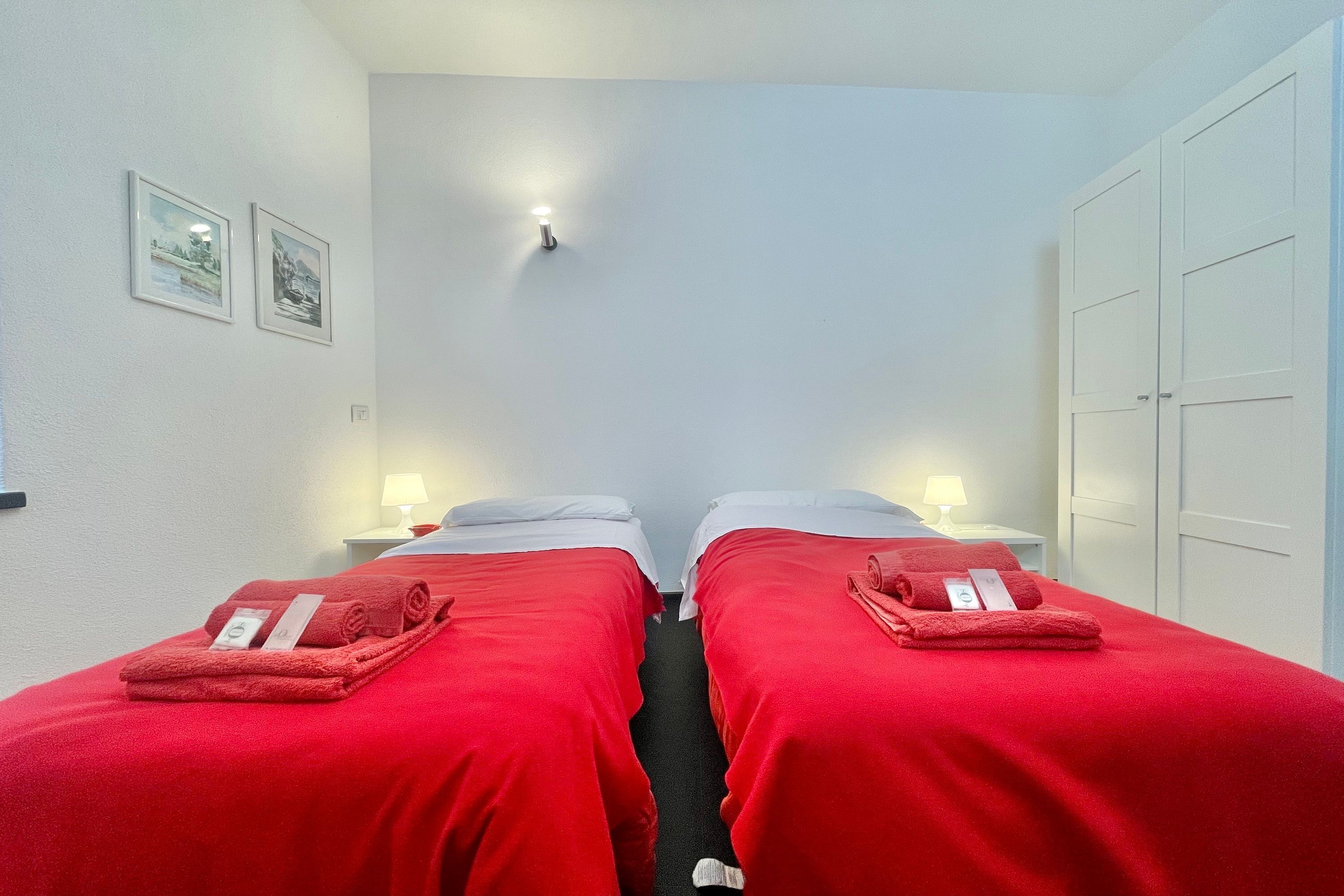Quarto de hotel com duas camas, cada uma com um cobertor vermelho e uma toalha. Há duas mesas de cabeceira com abajures e as paredes são brancas.