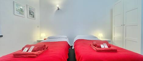 Quarto de hotel com duas camas, cada uma com um cobertor vermelho e uma toalha. Há duas mesas de cabeceira com abajures e as paredes são brancas.