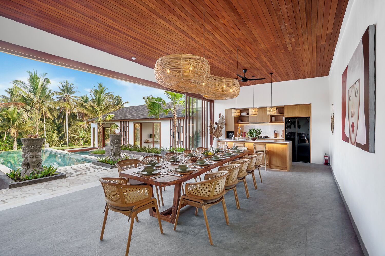 Uma sala de jantar moderna com uma mesa de madeira comprida e cadeiras de vime, uma cozinha com armários de madeira e vista para uma piscina e plantas tropicais.