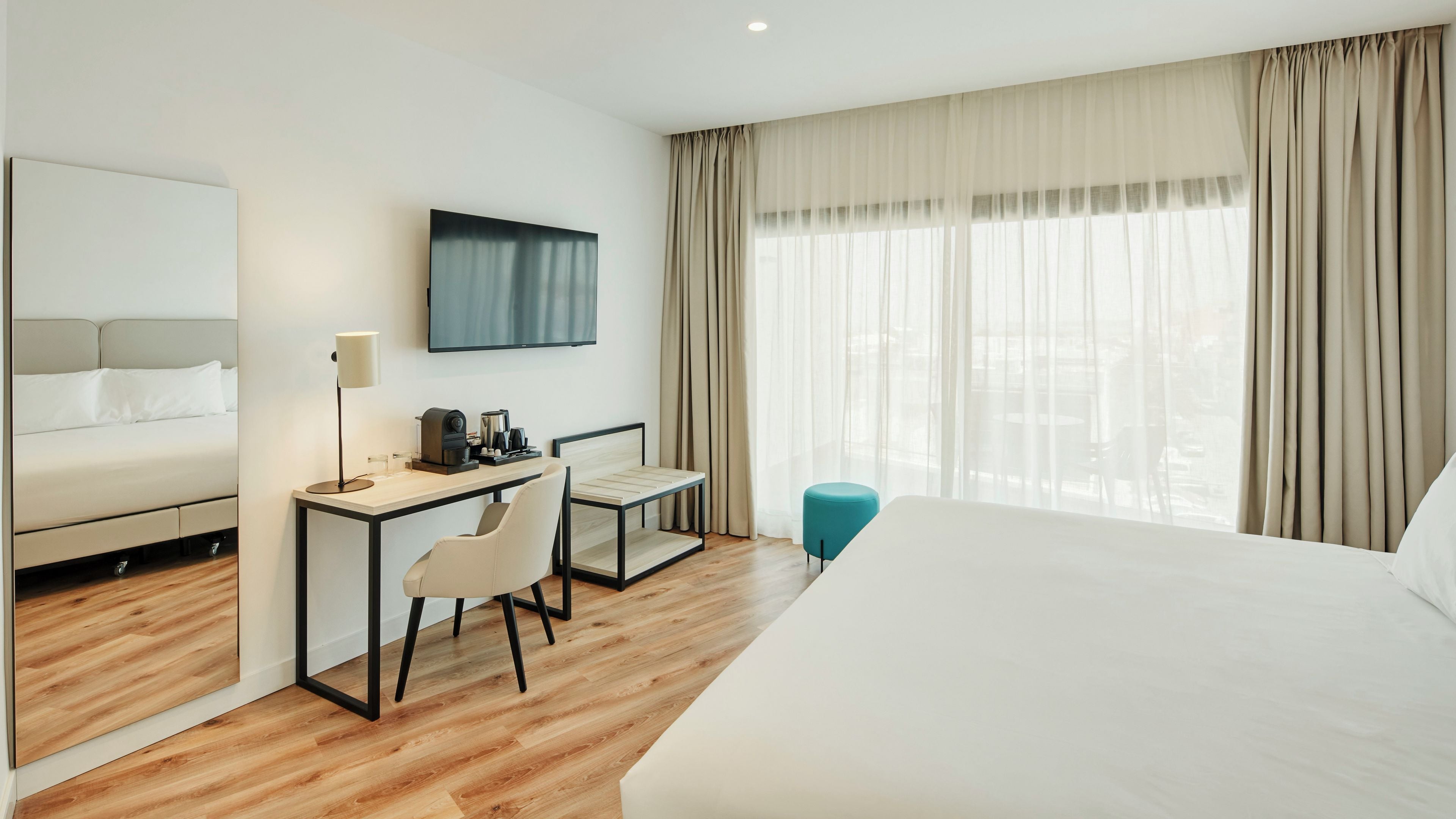 Quarto de hotel com cama, escrivaninha, cadeira, televisão e vista da cidade através de grandes janelas.