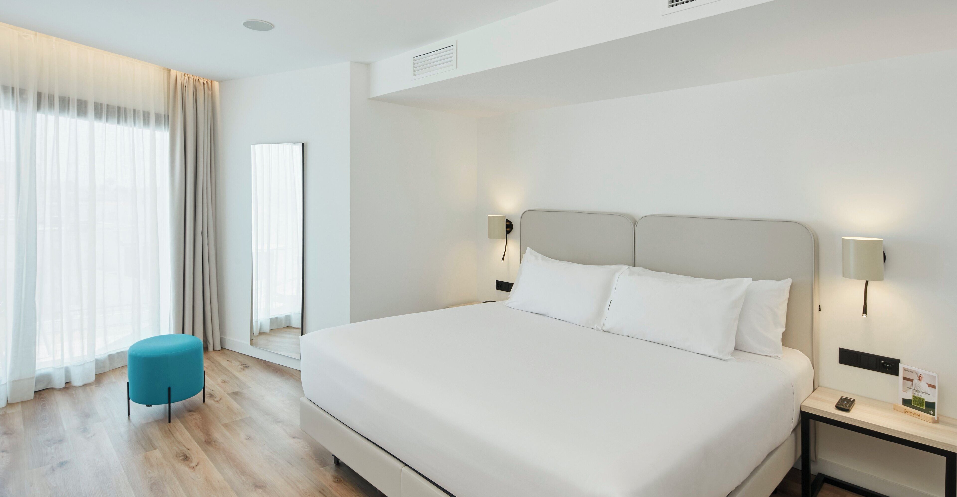 Quarto de hotel moderno com uma cama grande, duas mesas de cabeceira, uma mesinha de apoio, uma cadeira pequena azul e uma janela grande com cortinas finas.