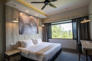 Quarto de hotel com cama, escrivaninha, cadeira, ventilador de teto e uma grande janela com cortinas.