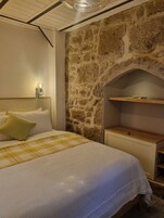 Quarto com parede de pedra, cama com manta quadriculada amarela e branca, mesa de cabeceira e luminária fixada na parede.