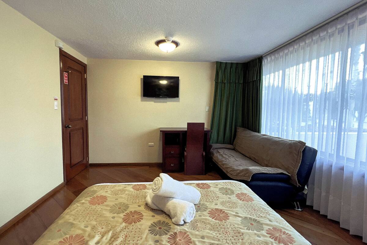 Quarto de hotel com cama, escrivaninha, cadeira, televisão e janela com cortinas.
