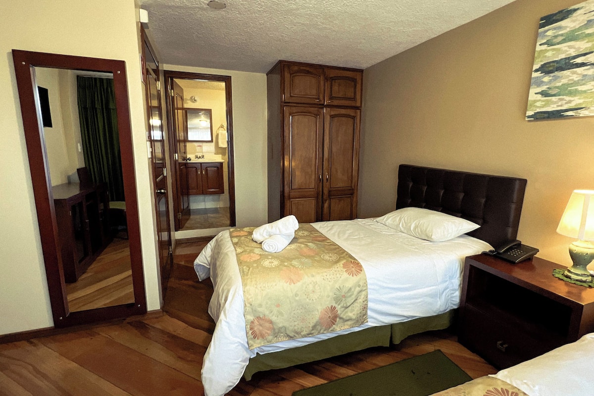Quarto de hotel com cama, mesa de cabeceira, luminária, espelho e guarda-roupa.
