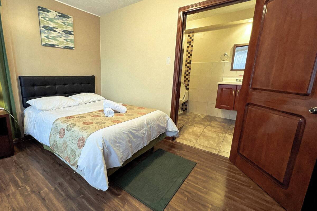 Quarto de hotel com cama, mesa de cabeceira, luminária, tapete verde, banheiro com vaso sanitário e espelho.