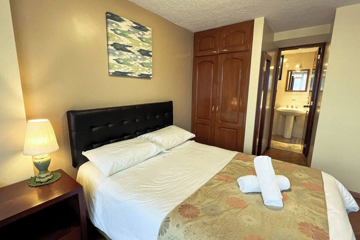 Quarto de hotel com uma cama, mesa de cabeceira com abajur, guarda-roupa de madeira e banheiro visível por uma porta aberta.