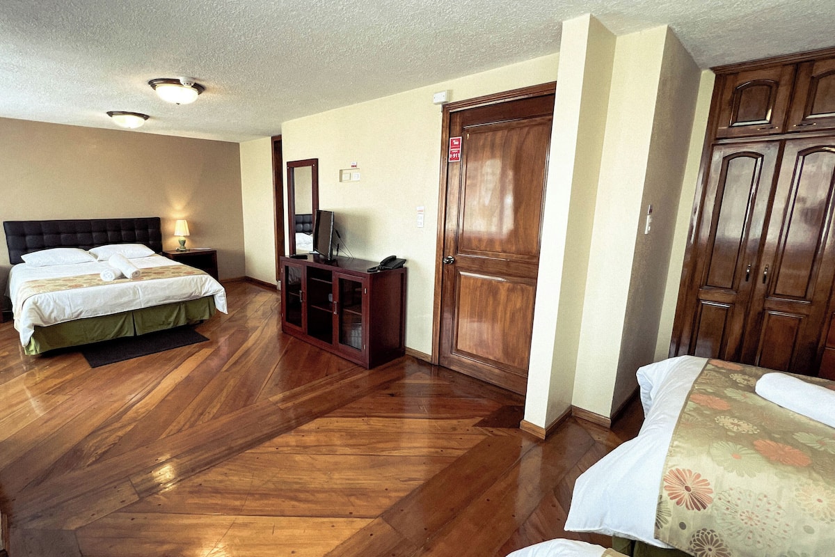 Quarto de hotel com piso de madeira, uma cama com encosto, mesa de cabeceira, escrivaninha com computador, guarda-roupa e uma porta.