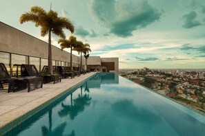 Piscina infinita no terraço com espreguiçadeiras e palmeiras, com vista para a cidade.