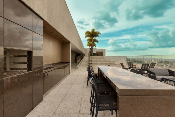 Área de cozinha externa com bancada comprida e banquetas, uma palmeira e vista para a cidade.