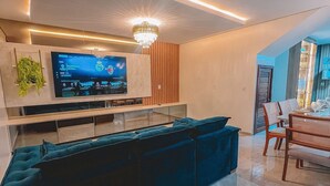 Uma sala de estar moderna com uma televisão de tela grande, um sofá azul escuro, uma mesa de jantar de madeira com cadeiras e um lustre.