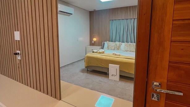 Quarto de hotel com parede de painel de madeira, cama com colcha amarela, mesa de cabeceira e criado-mudo.