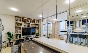 Uma sala de estar moderna com uma televisão de tela plana grande, nichos com objetos decorativos, um espaço para refeições com bar, e uma janela ampla com cortinas.