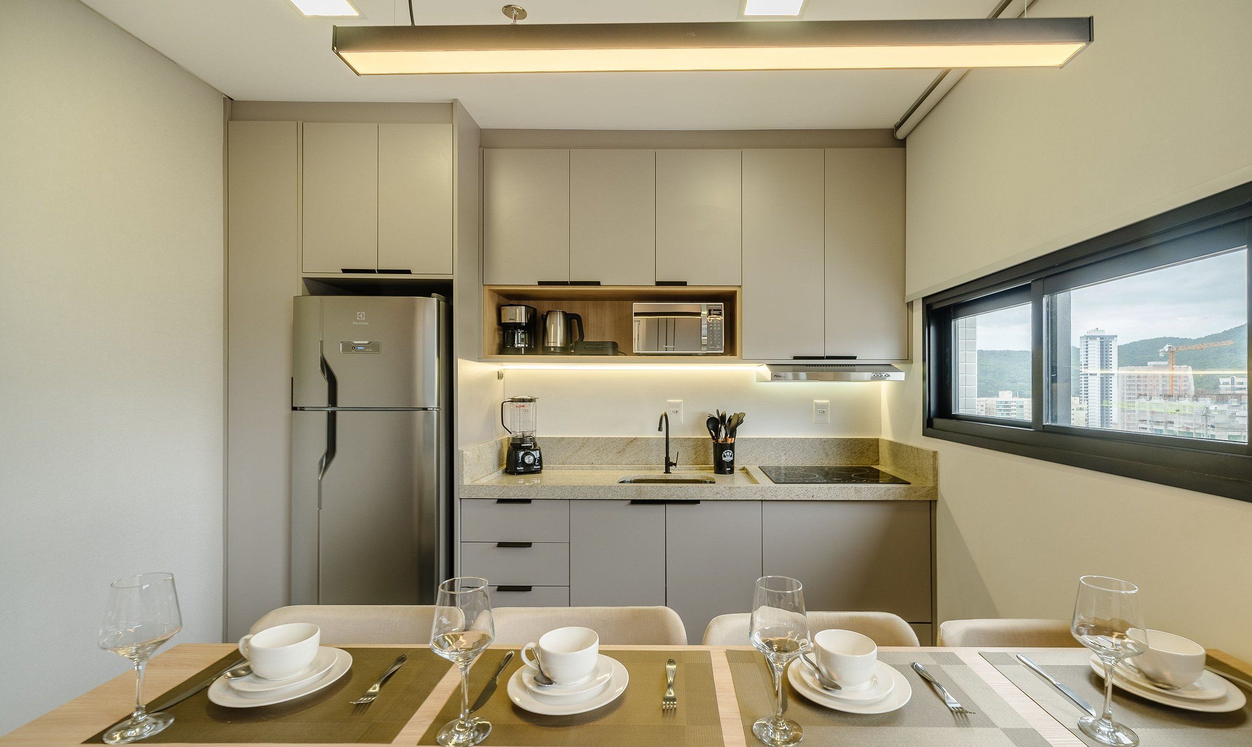 Uma cozinha moderna com eletrodomésticos embutidos, área de jantar com mesa posta para quatro pessoas e vista da cidade pela janela.