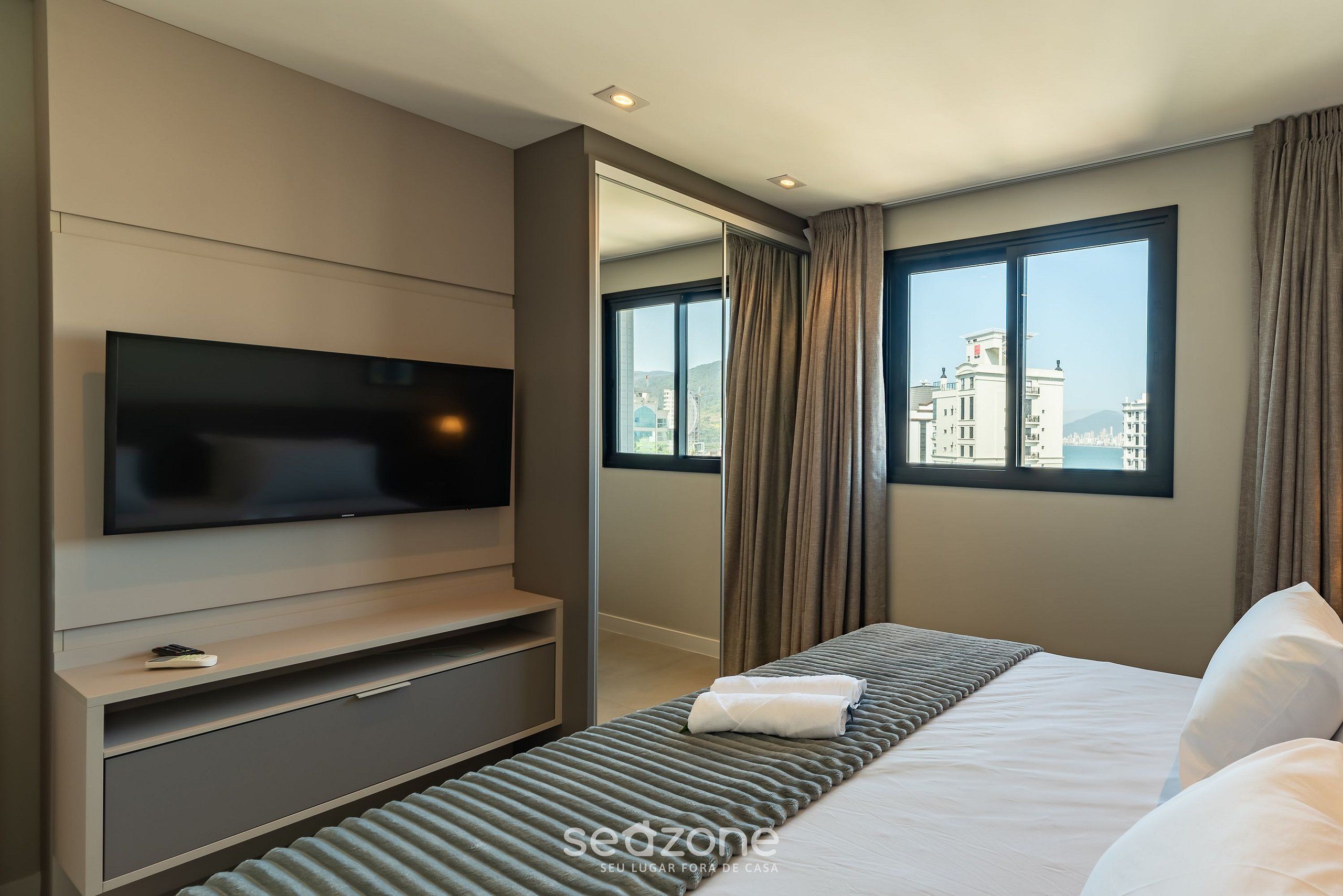Quarto de hotel moderno com uma grande TV de tela plana, uma cama com lençóis brancos, uma mesa de cabeceira e vista para edifícios pela janela.