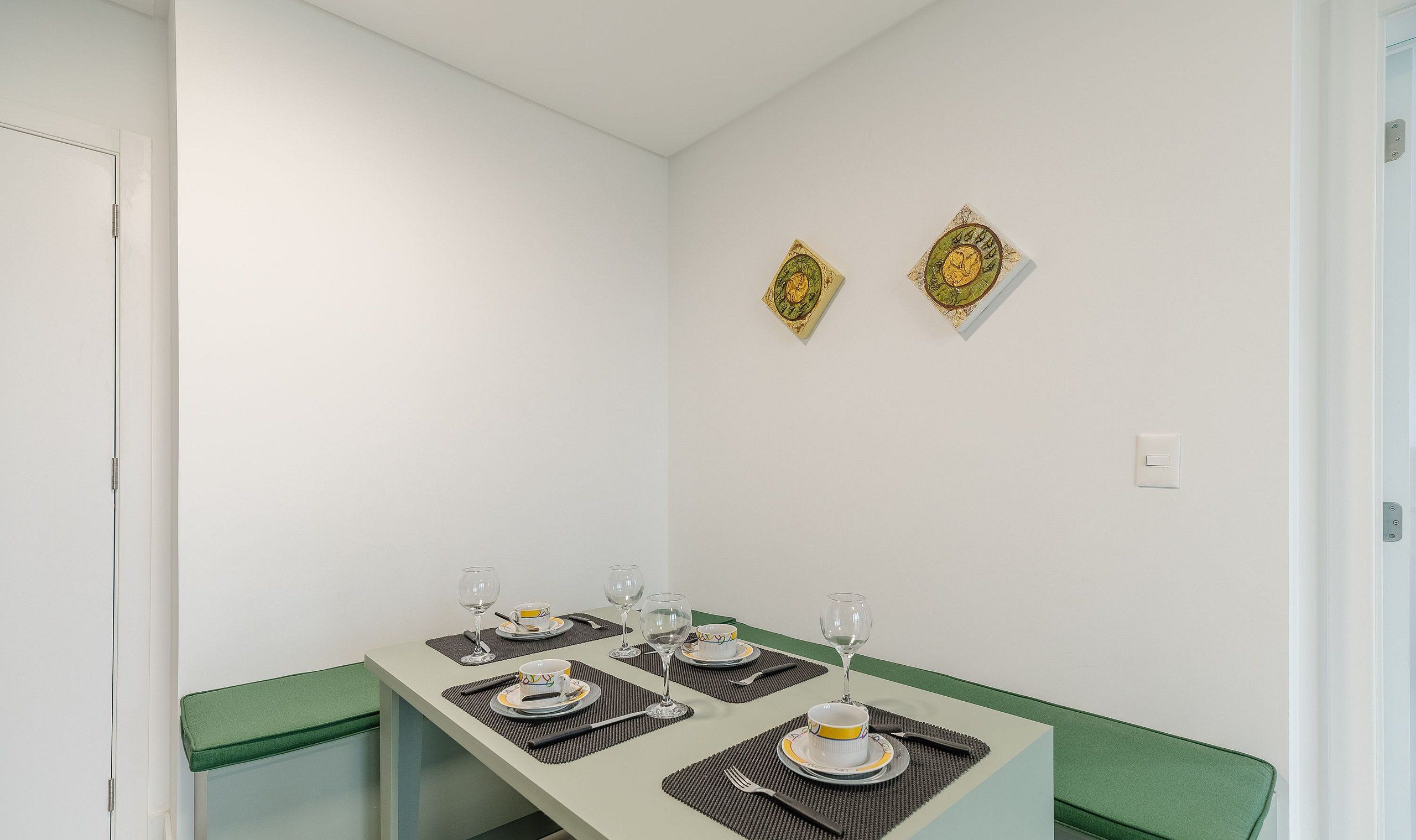 Uma área de refeições com uma mesa posta para quatro pessoas, incluindo pratos, talheres e copos.