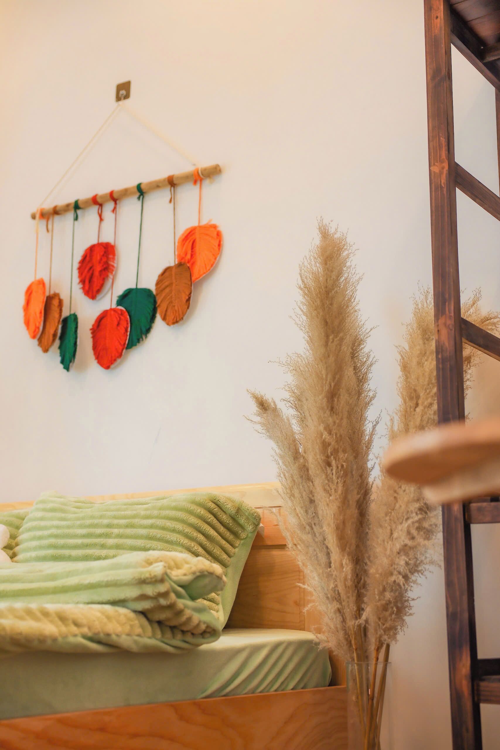 Uma cama com manta e almofadas verdes, um ornamento fixado na parede com folhas coloridas e capim-do-campo em um vaso.
