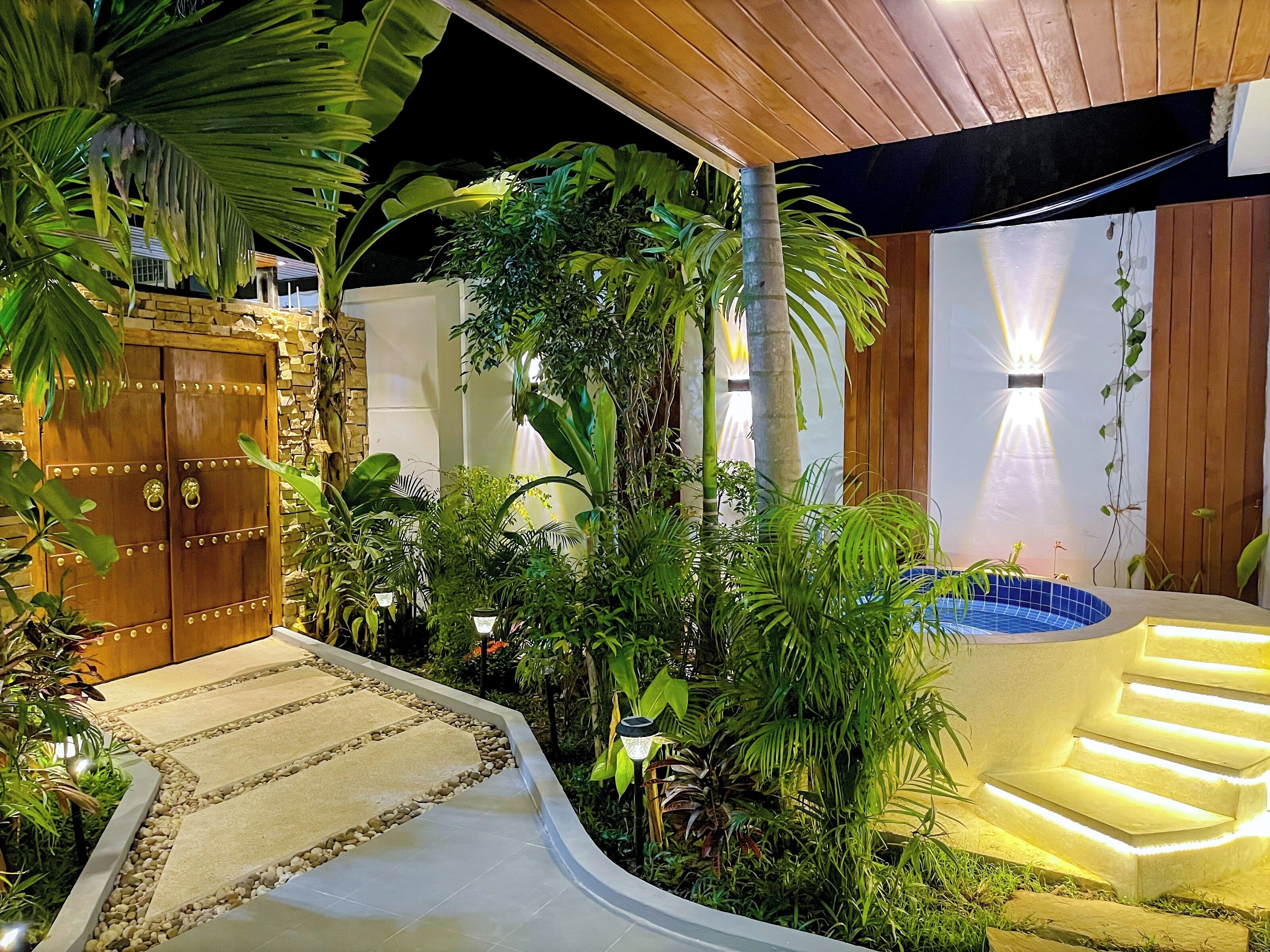 Área externa bem iluminada com porta de madeira, caminho sinuoso e piscina com degraus iluminados.