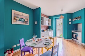 Uma sala de jantar com paredes verde-água, uma mesa branca e cadeiras roxas. Cozinha visível através de uma porta aberta.