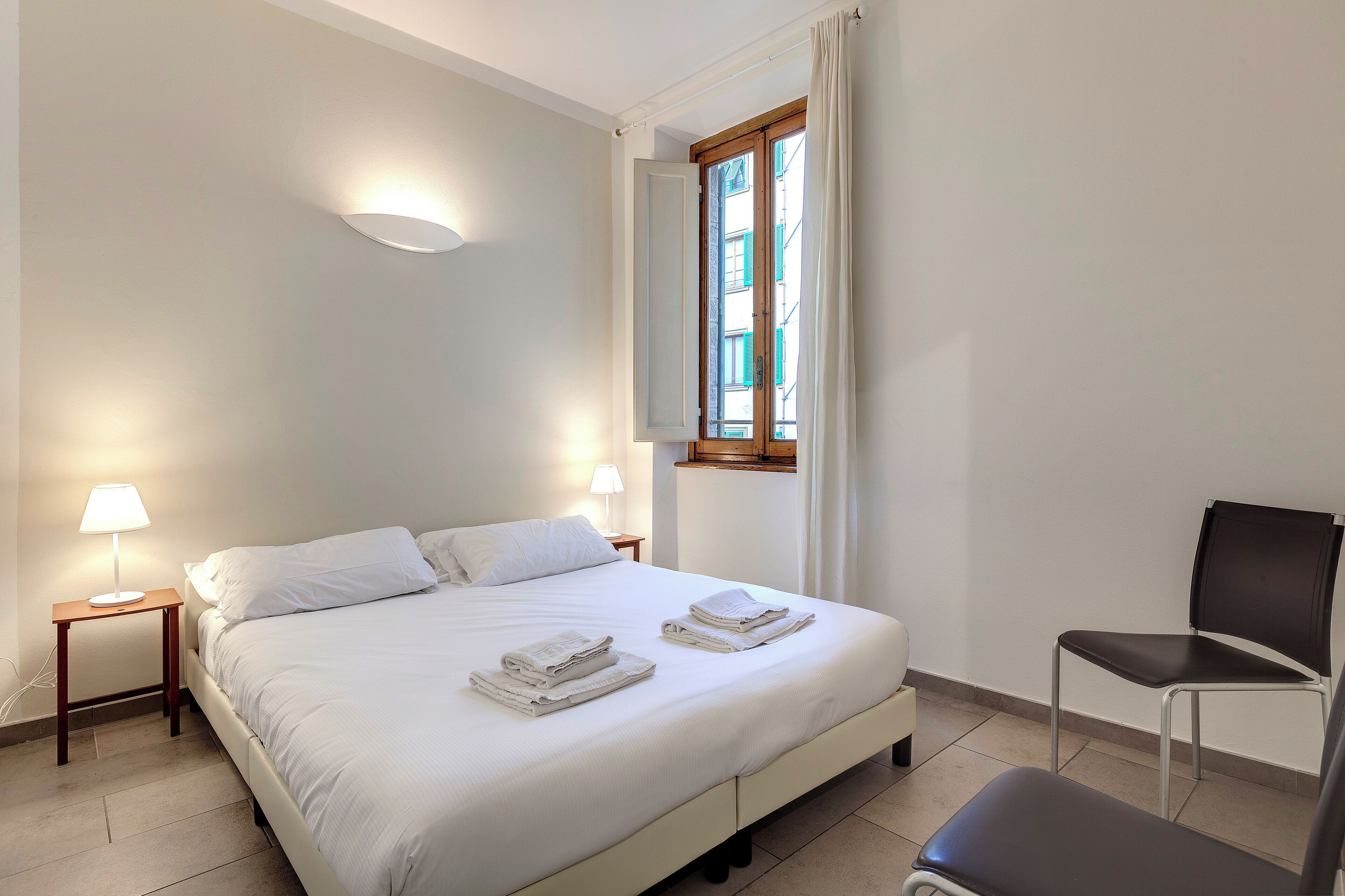 Cama bem arrumada com lençóis brancos e duas toalhas dobradas, uma luminária de cabeceira, uma cadeira e uma janela com cortinas.