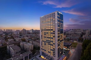 Um edifício alto e moderno com a inscrição "ASCOTT" na fachada, cercado por uma área urbana densa e diversos outros edifícios.