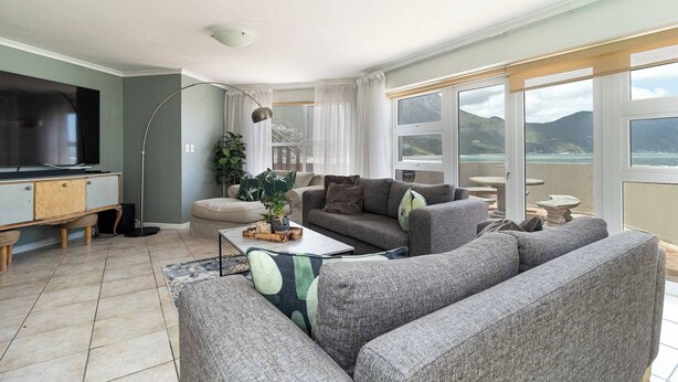 Uma sala de estar moderna com um conjunto de sofá, uma mesa de centro e uma televisão de tela plana. Há uma varanda com vista para as montanhas.