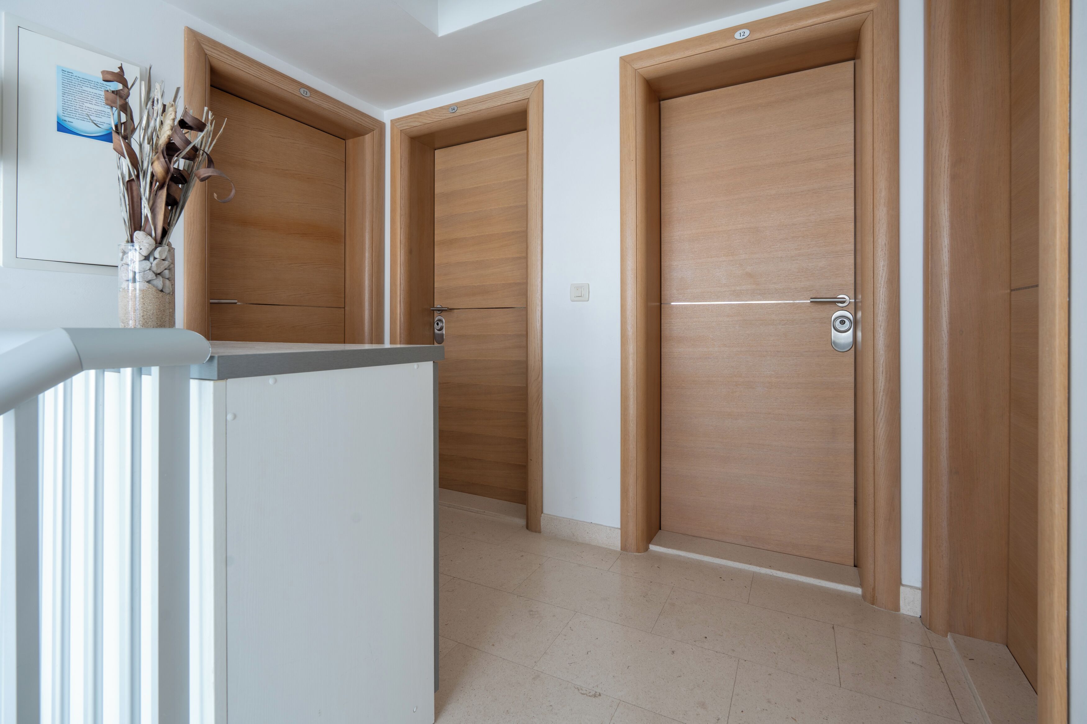 Corredor com portas de madeira, um balcão de recepção branco e um vaso com flores secas.