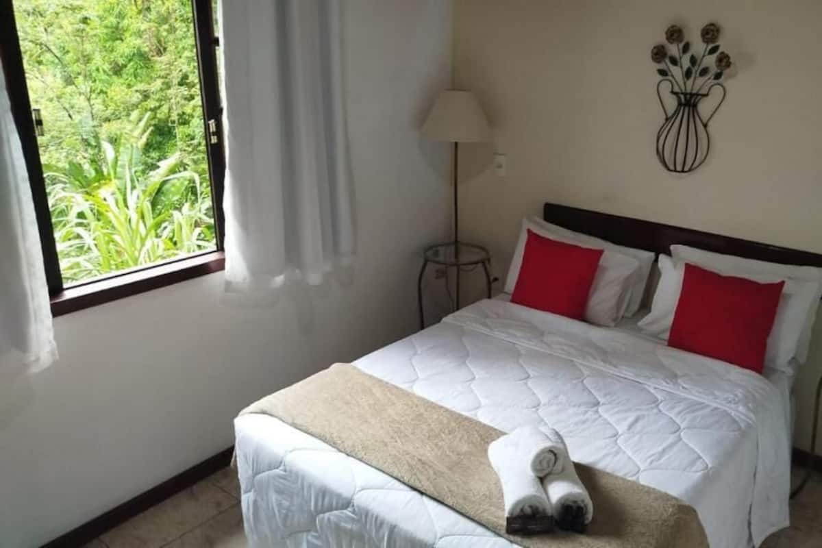 Cama bem arrumada com travesseiros brancos e vermelhos, um cobertor bege e uma toalha enrolada sobre uma mesinha.